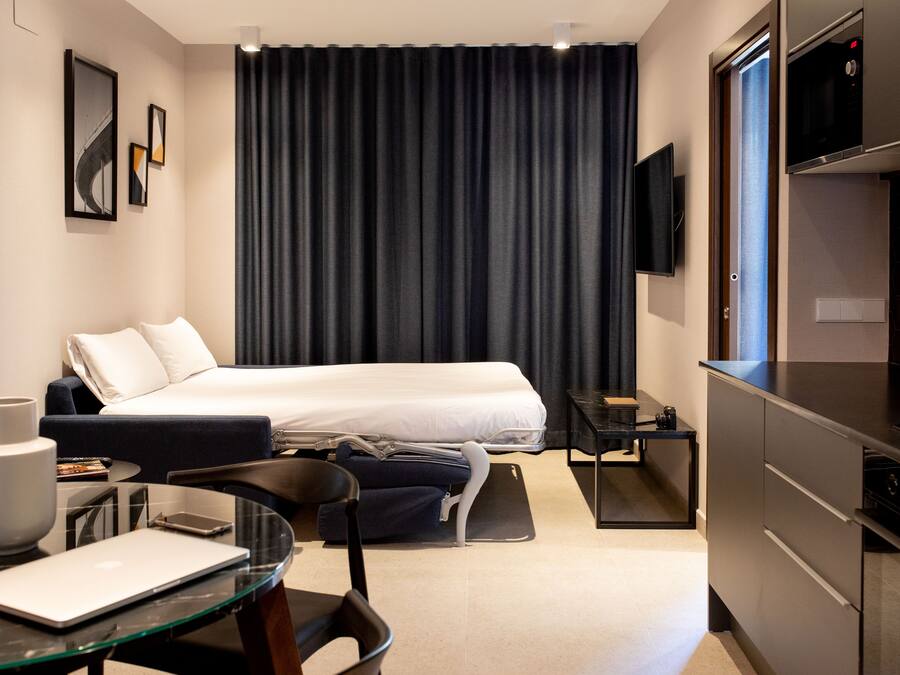 Quarto de hotel moderno com uma cama bem arrumada, uma mesa de vidro com um laptop, uma cadeira, uma pequena escrivaninha com uma lâmpada e uma área de cozinha com micro-ondas.