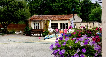 Uma casa rústica com telhado de telhas, quintal cercado e jardim com flores roxas.