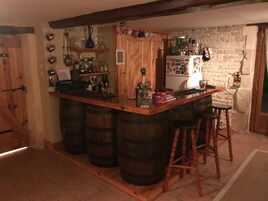 Uma área de bar rústica com barris de madeira como bancos, um balcão de madeira e diversos frascos e copos.