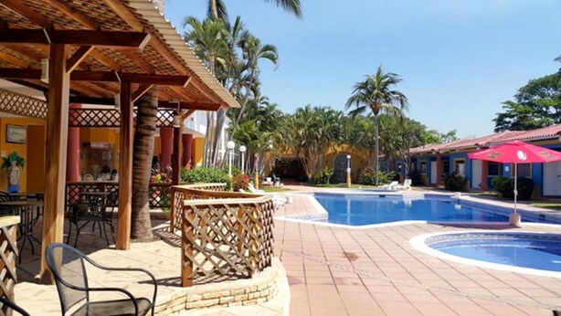 Área da piscina com espaço coberto para relaxar, palmeiras e céu azul límpido.