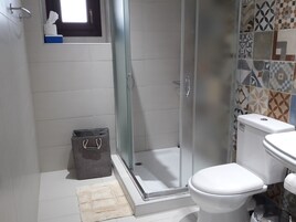 Uma casa de banho com chuveiro em cabine de vidro, sanita branca e parede revestida a mosaicos com padrão geométrico.
