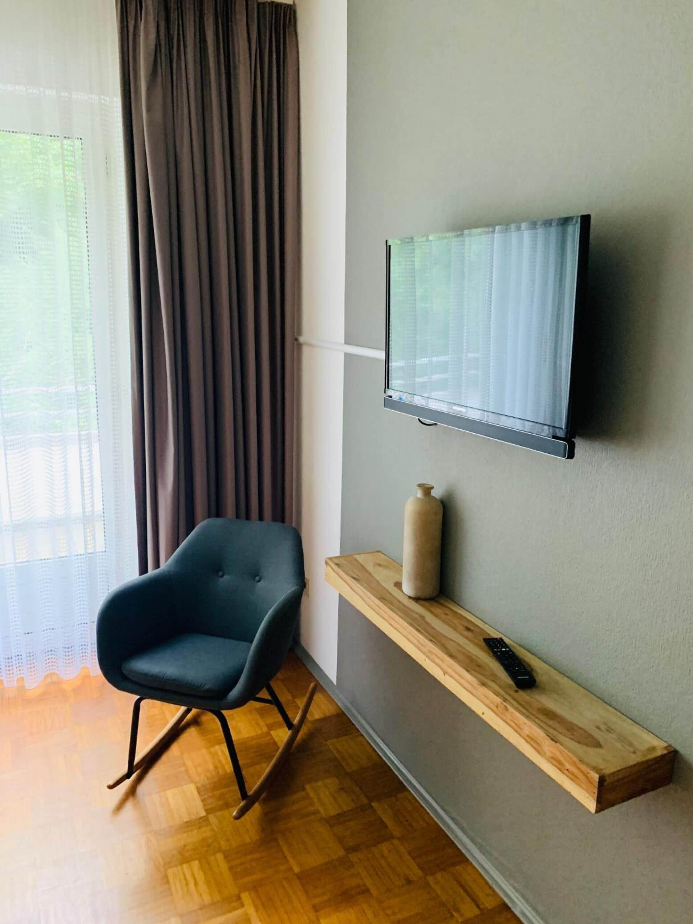 Quarto moderno com piso de madeira, uma cadeira azul, uma TV fixada na parede e uma prateleira de madeira com um vaso e um controle remoto.