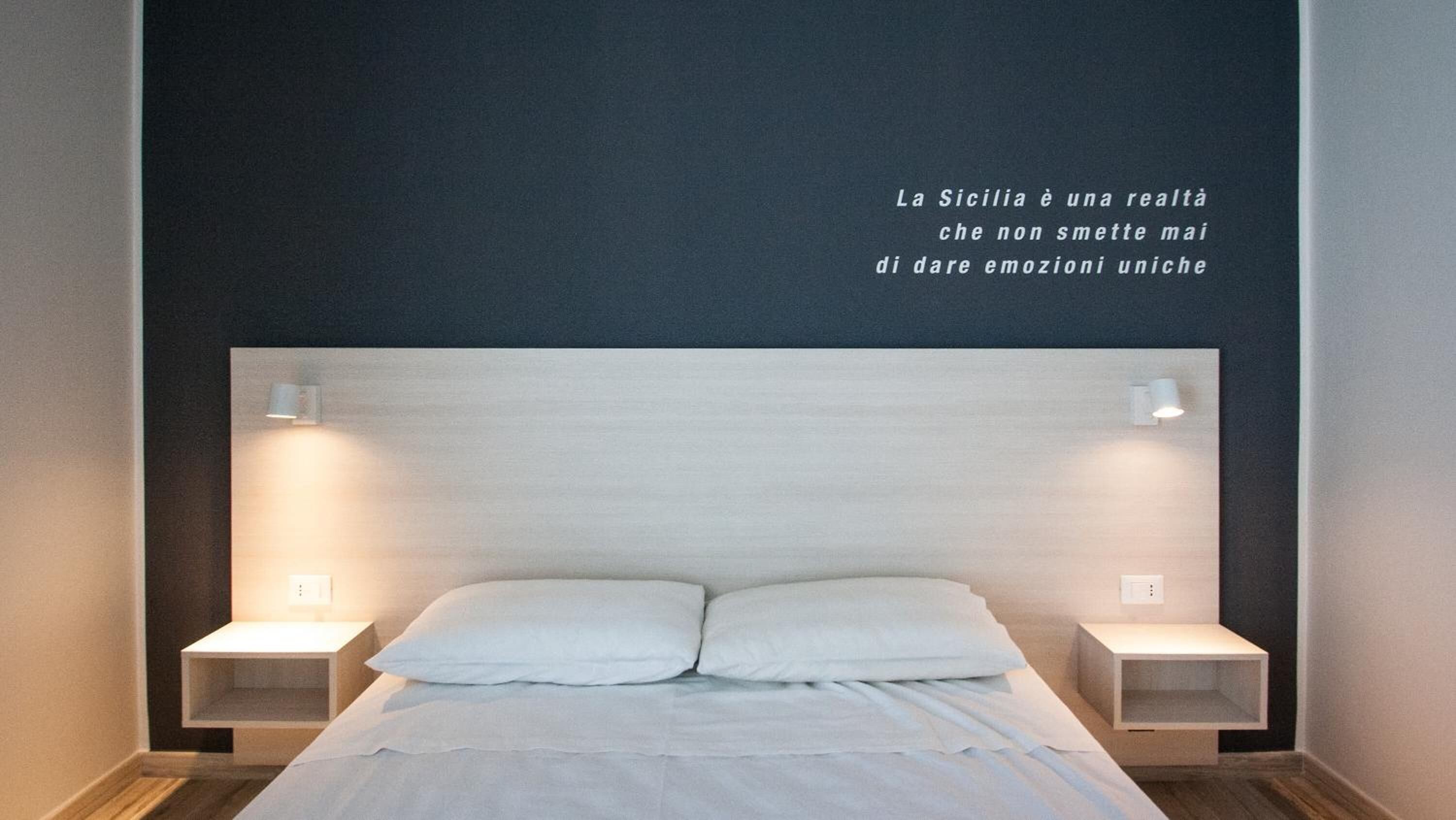 Quarto moderno com uma cama grande, duas mesas de cabeceira e uma parede escura com uma citação em italiano.