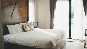 Uma cama grande com cabeceira de madeira, uma mesinha de cabeceira e vista para o exterior através de uma porta de vidro deslizante.