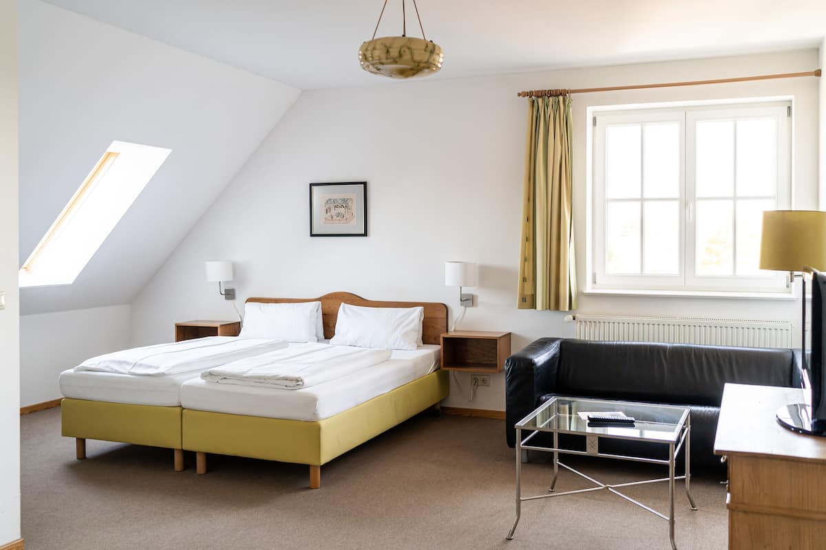 Quarto de hotel com cama, sofá de couro preto, mesa de centro de vidro, escrivaninha com abajur, janela com cortinas, quadro na parede e luminária no teto.