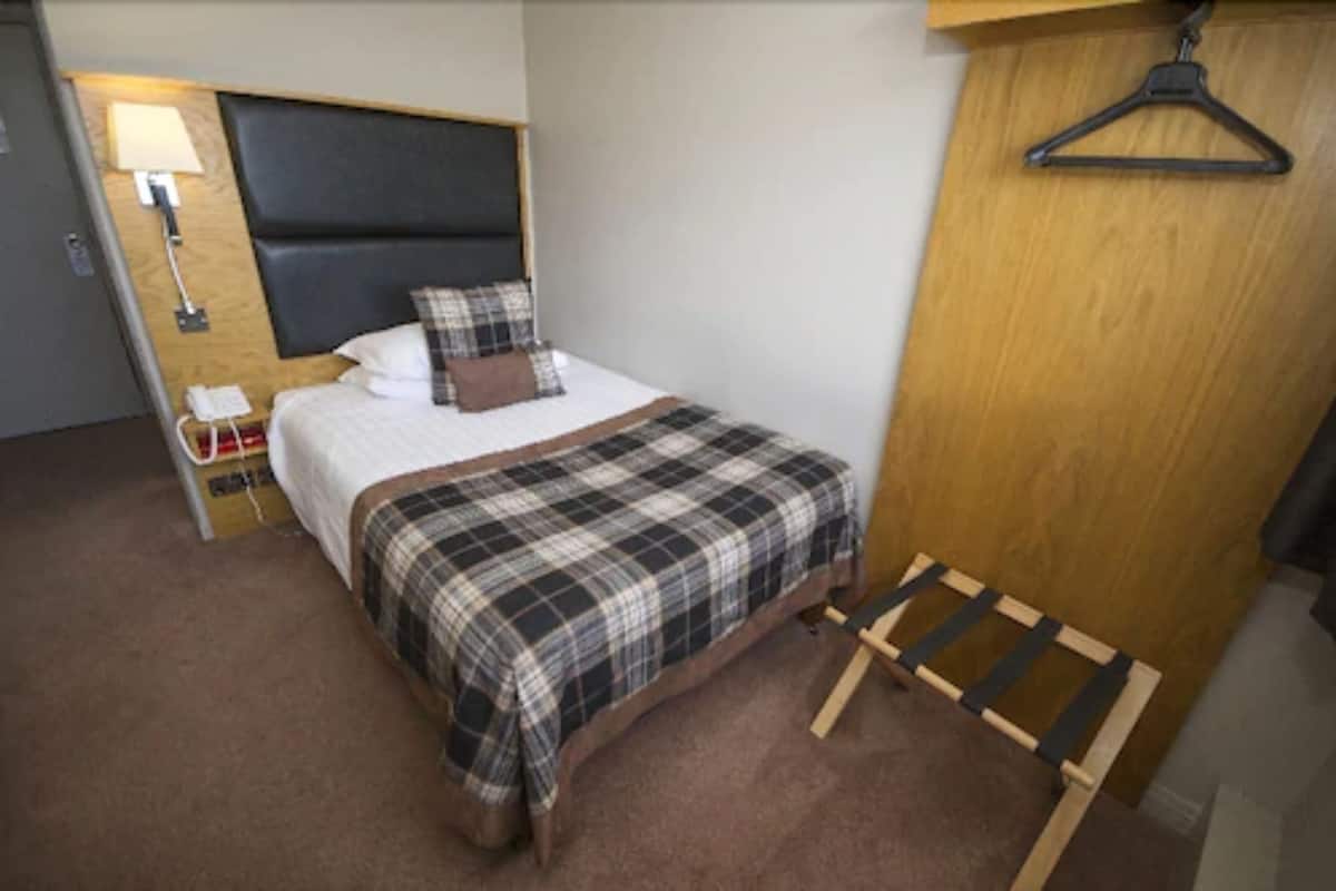 Quarto com cama, mesa de cabeceira com abajur, cabideiro e escada de madeira.