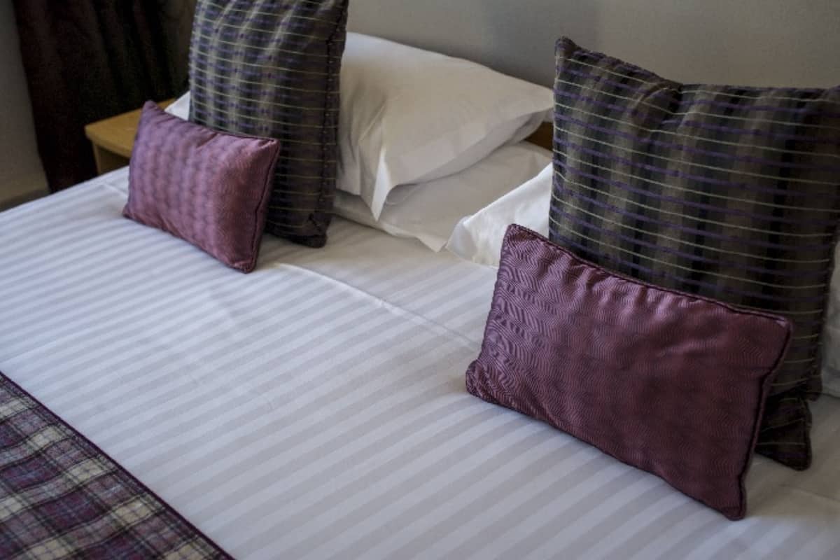 Cama bem arrumada com roupa de cama listrada e duas almofadas, uma roxa e outra xadrez, em quarto de hotel.