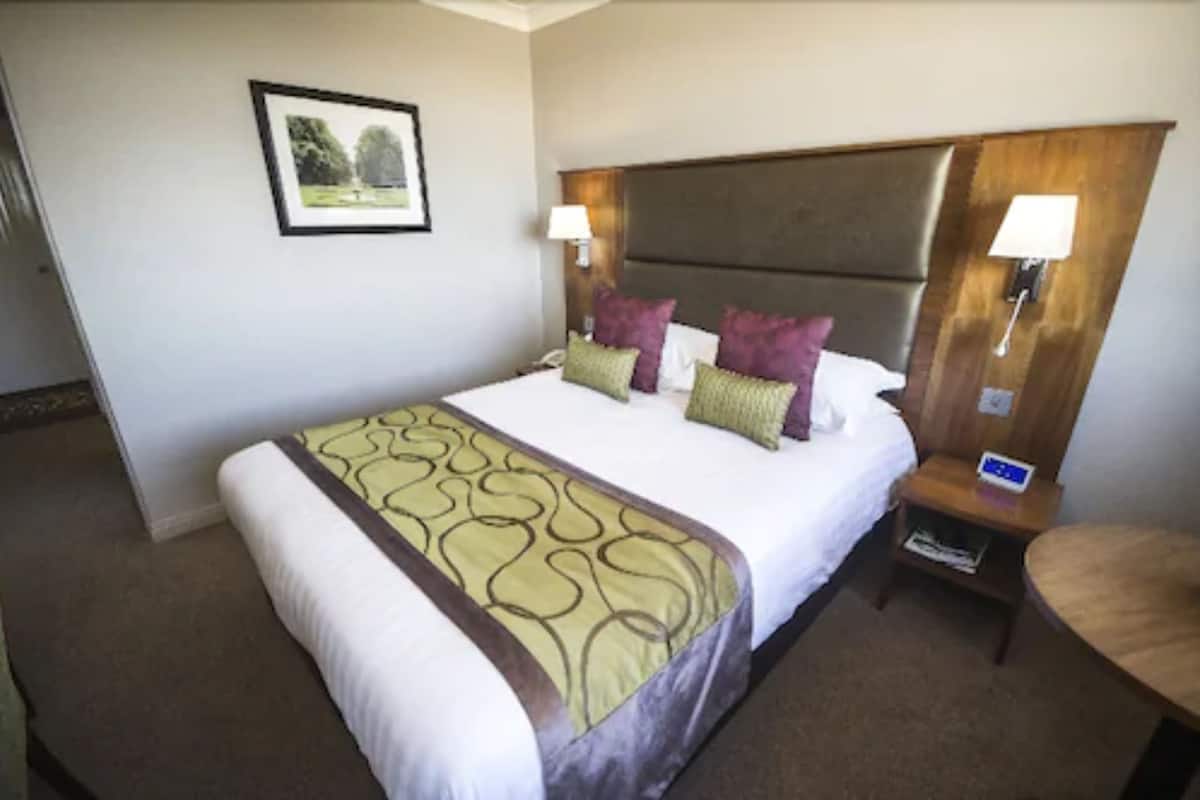 Quarto de hotel com uma cama grande, duas mesas de cabeceira com abajures, um quadro na parede e piso coberto por carpete.