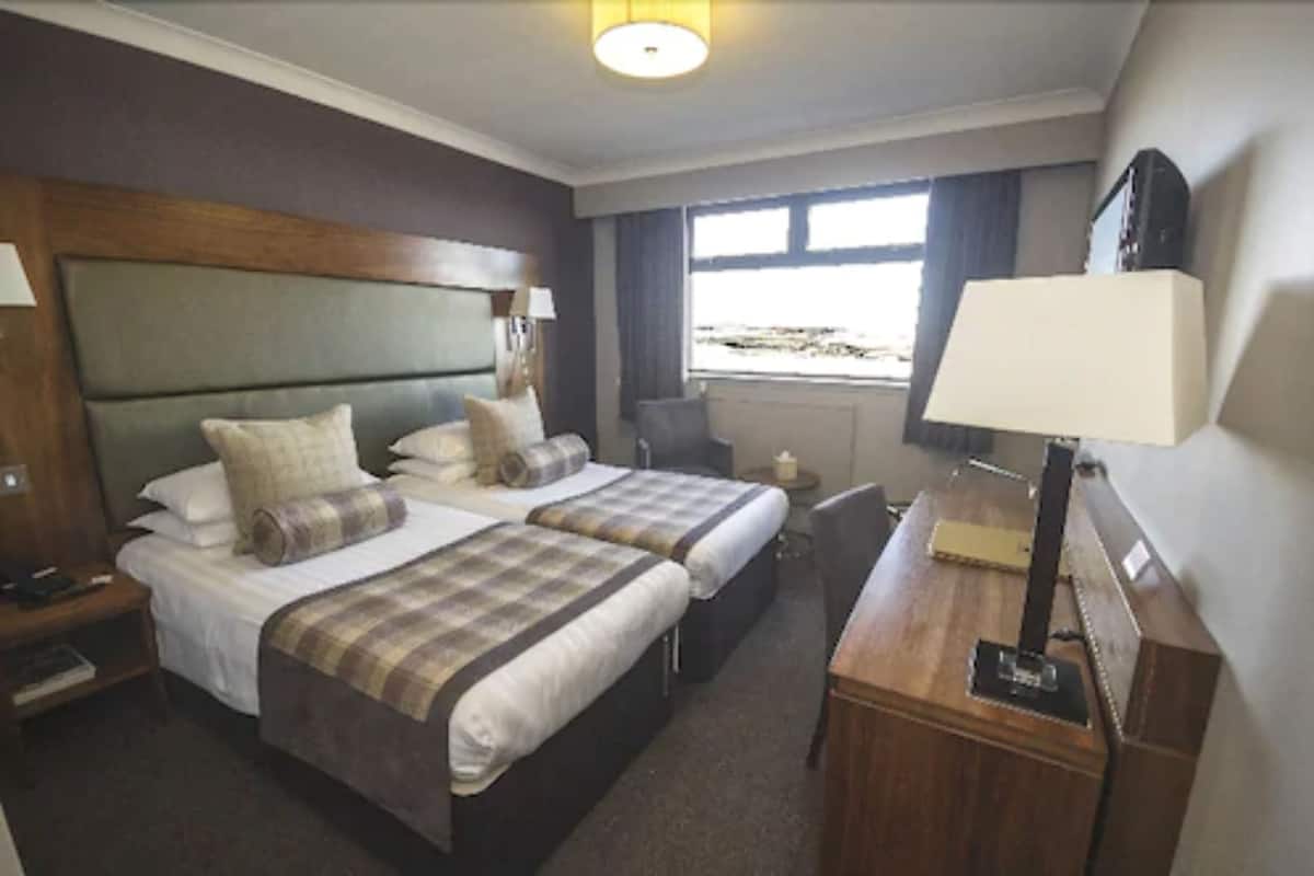 Quarto de hotel com duas camas, uma escrivaninha, uma cadeira e uma janela com vista.