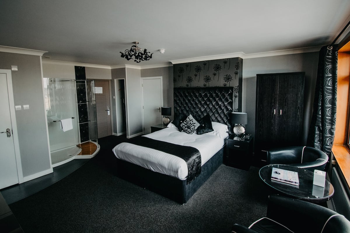 Quarto de hotel moderno com uma cama grande, box de chuveiro de vidro, cabeceira escura, poltrona de couro preta e uma mesinha de cabeceira com um livro.