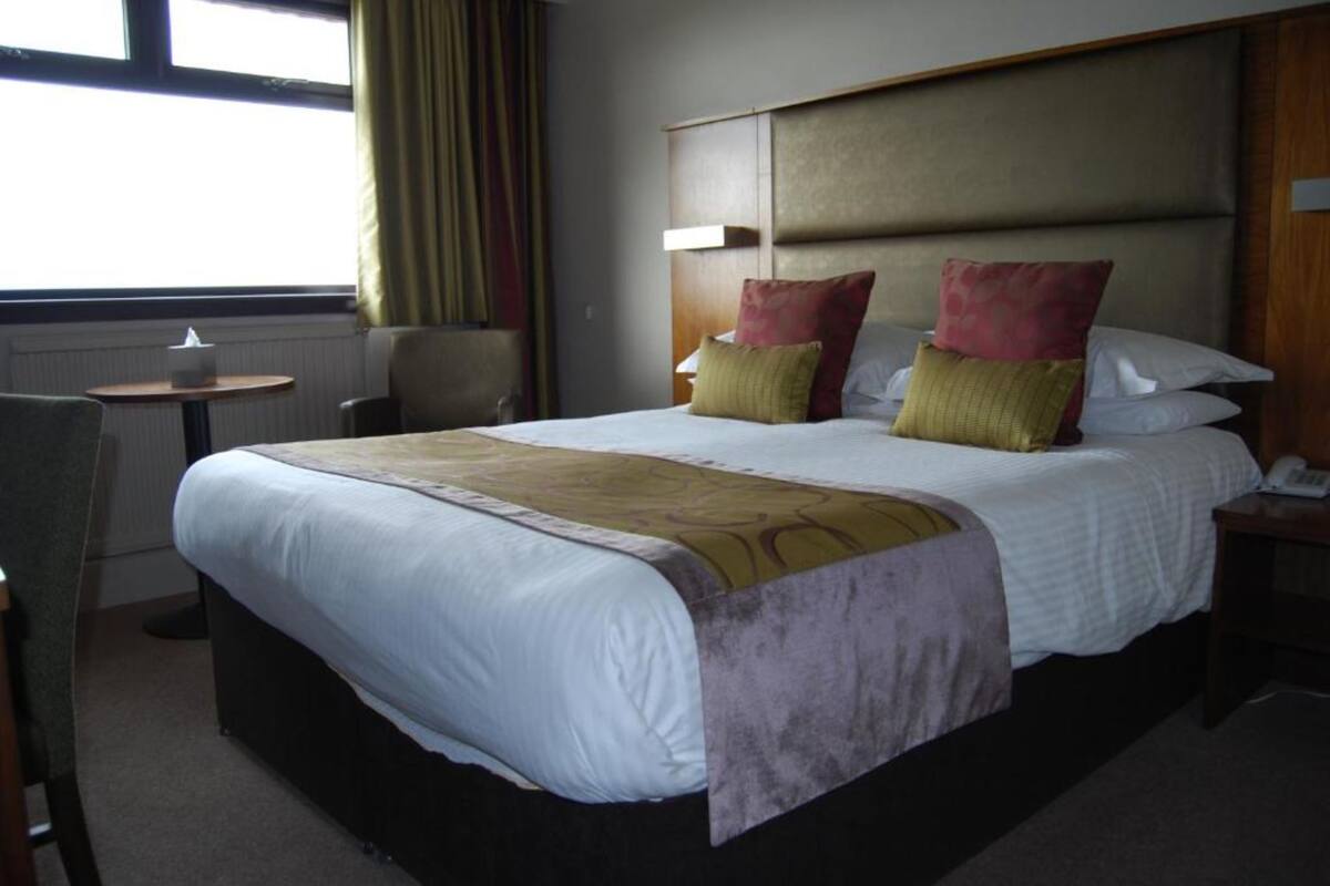 Quarto de hotel com uma cama grande, mesa de cabeceira, uma cadeira e janela com cortinas.