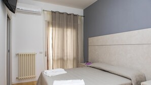 Quarto com cama, radiador, TV fixada na parede e janela com cortinas.