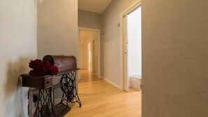 Um corredor com piso de madeira, uma mesa de costura antiga com uma bolsa de couro e rosas vermelhas, e uma porta no final.