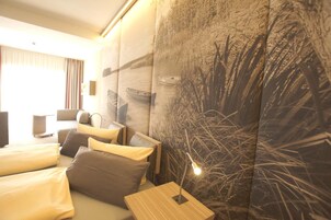 Quarto de hotel com uma cama grande, uma mesa de cabeceira com abajur e um mural com tema de natureza na parede.