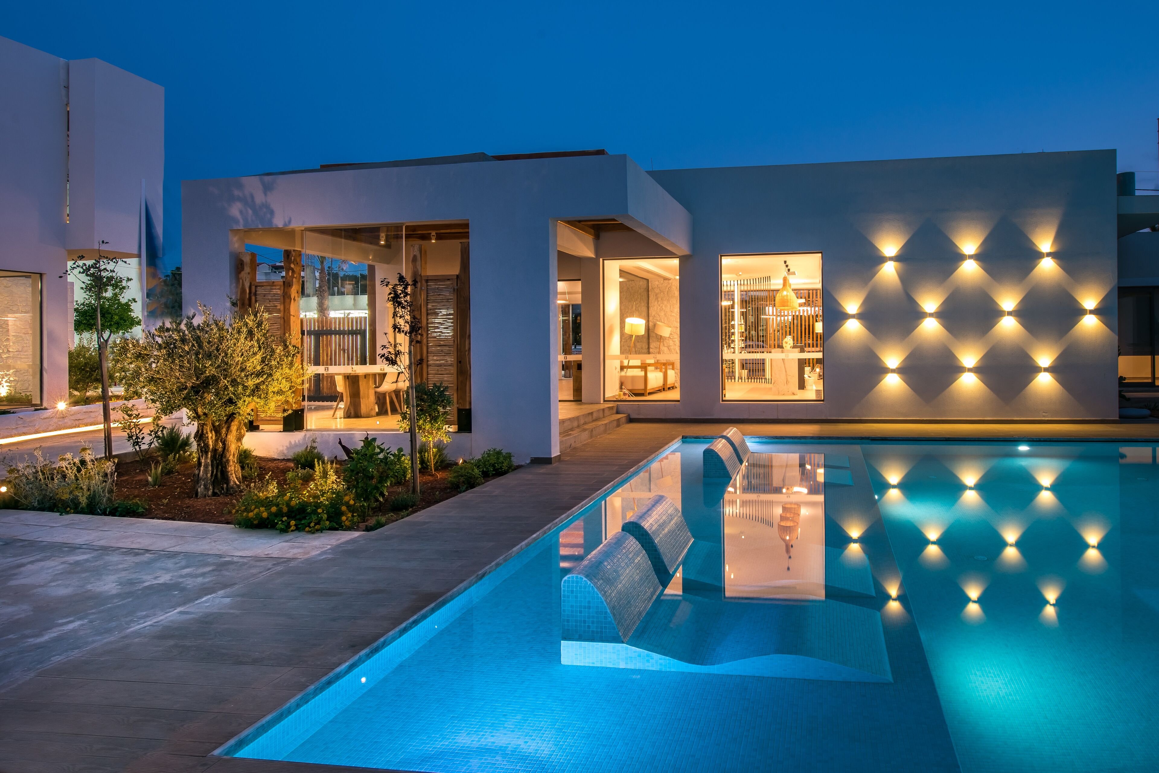Uma área de piscina moderna, com piscina transparente, adjacente a um edifício com grandes janelas e um padrão de luz geométrica na fachada.