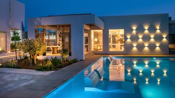 Uma área de piscina moderna, com piscina transparente, adjacente a um edifício com grandes janelas e um padrão de luz geométrica na fachada.