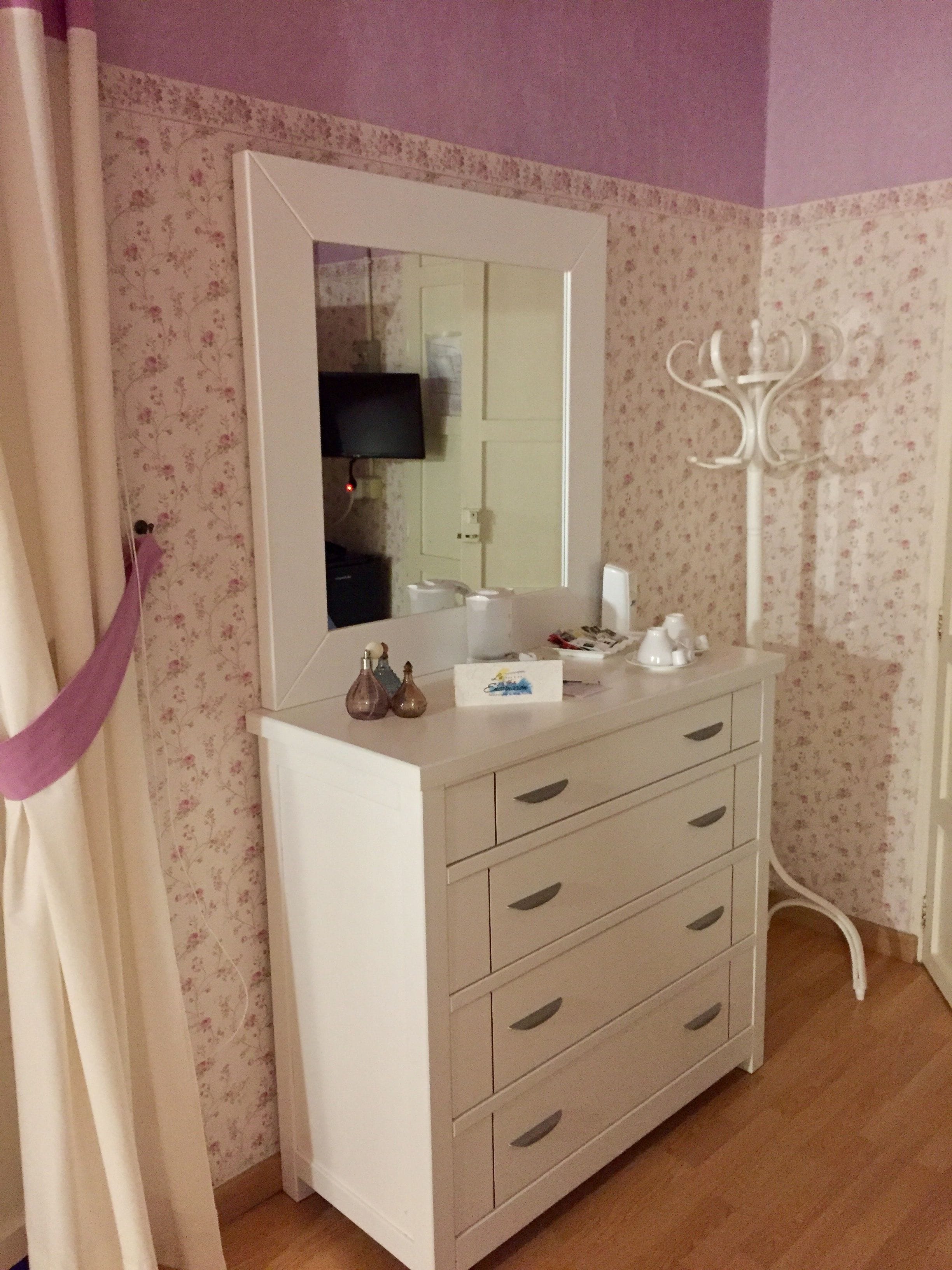 Quarto com cômoda branca, espelho, televisão de tela plana e papel de parede estampado com flores.