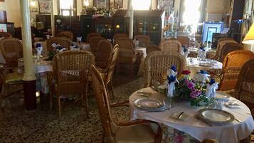 Restaurante com cadeiras e mesas de vime dispostas para refeições, com centros de mesa florais e chão de azulejos.