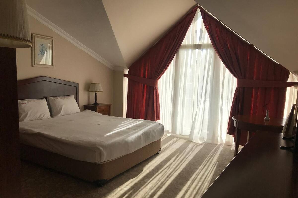 Quarto de hotel com uma cama grande, uma escrivaninha e uma janela com cortinas vermelhas.