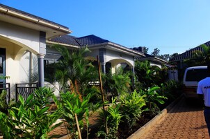 Um bairro residencial com um caminho de acesso pavimentado, uma van branca e diversas plantas tropicais.