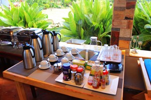 Disposição para o pequeno-almoço com máquinas de café, chávenas e acompanhamentos sobre uma mesa com vista para vegetação exuberante.