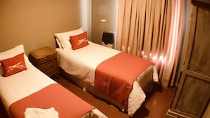 Quarto de hotel com duas camas, cada uma com colcha vermelha e branca, uma mesa de cabeceira de madeira e uma janela grande com cortinas.