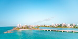 Paisagem costeira com um horizonte urbano, uma ponte ligando duas áreas terrestres, águas azuis cristalinas e diversos edifícios, incluindo apartamentos e hotéis.