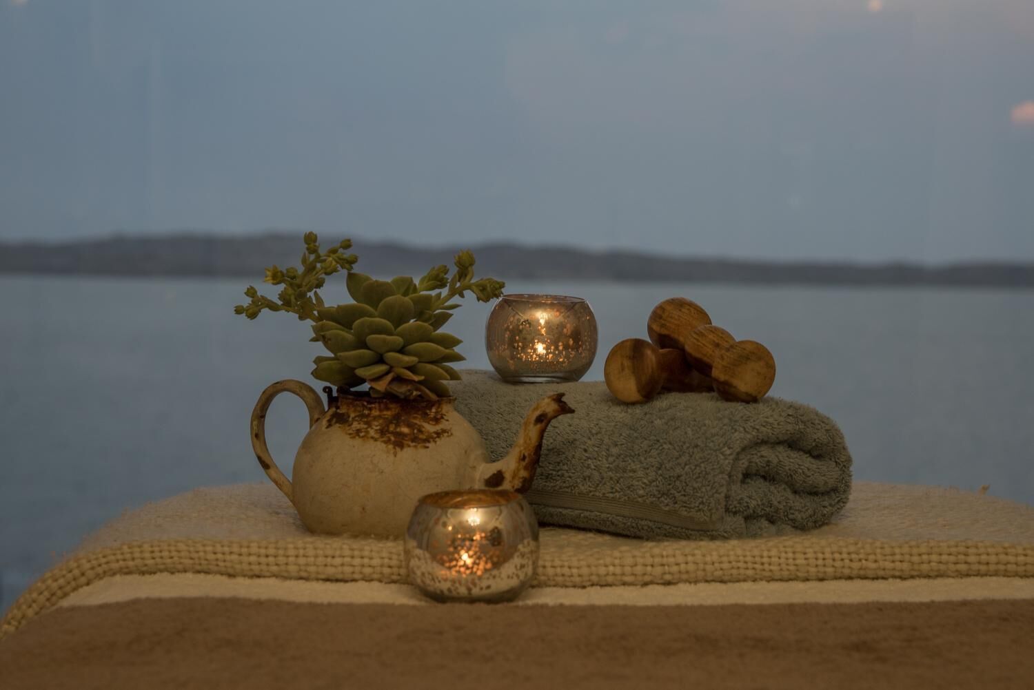 Um spa tranquilo com um bule de metal, uma toalha enrolada, bolas de madeira e uma planta suculenta sobre uma superfície texturizada, próximo a um corpo d'água calmo.