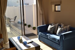 Uma sala de estar com um sofá azul, uma mesa de centro e vista para uma cerca branca e móveis de pátio.