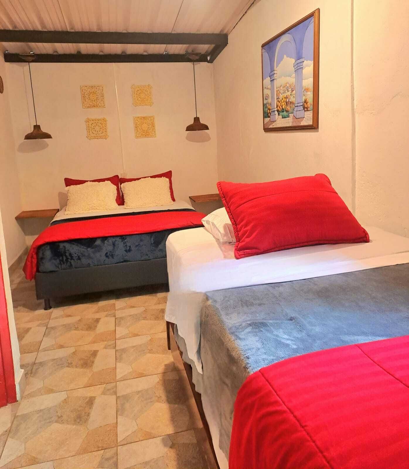 Quarto com duas camas, cada uma com roupa de cama vermelha. Há painéis de parede estampados e uma pintura acima das camas. O quarto possui um chão de azulejos e teto de madeira.