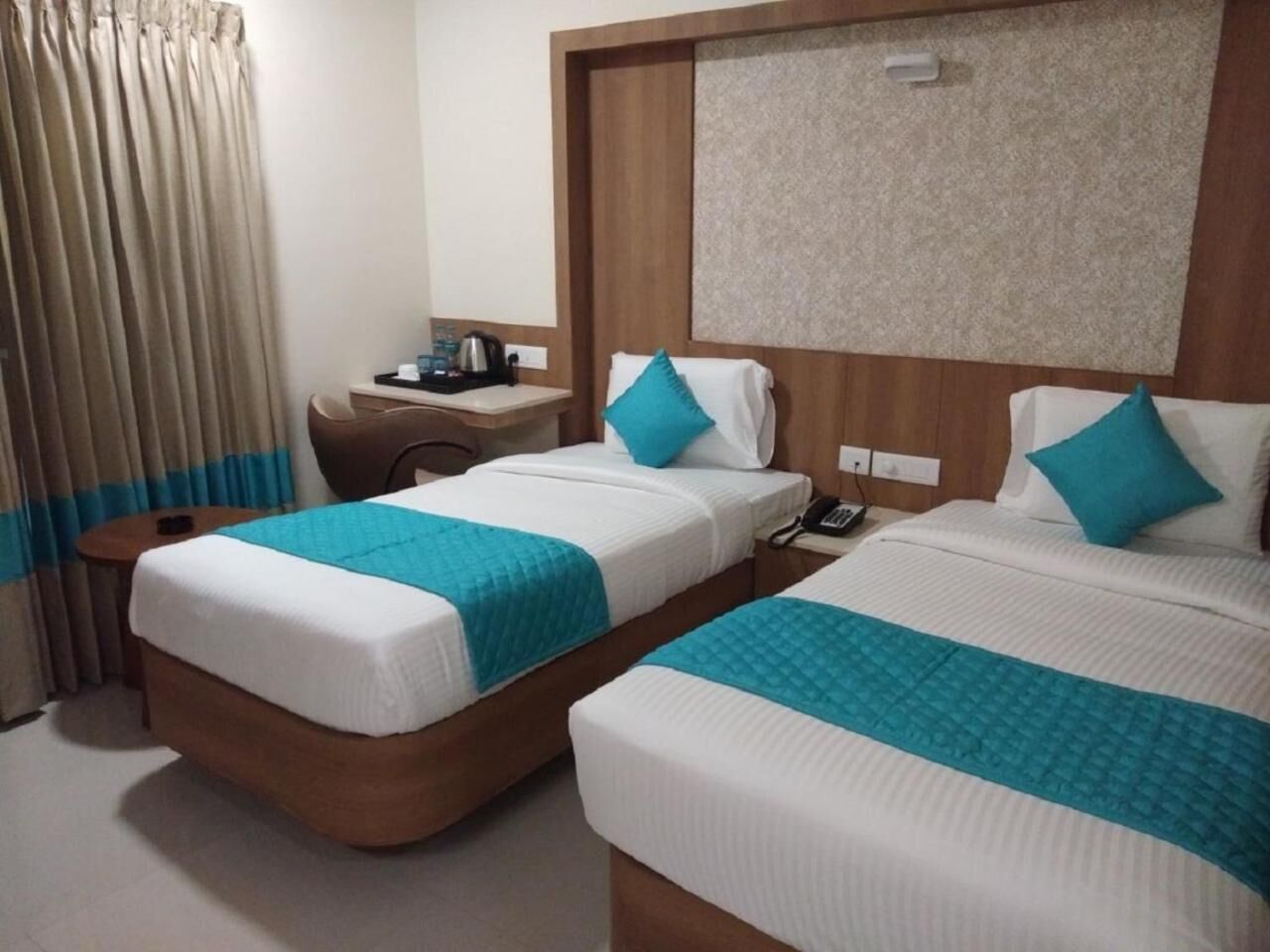 Quarto de hotel com duas camas, uma escrivaninha, uma cadeira, telefone e janela com cortinas.