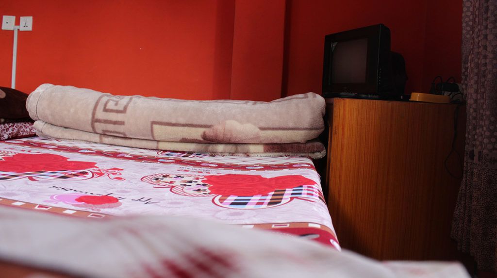 Uma cama com colcha estampada em vermelho e branco, um cobertor dobrado e uma mesa de cabeceira de madeira com uma televisão.