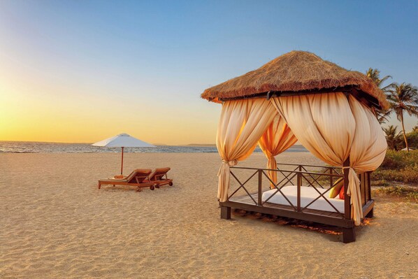 Uma cabana de praia com telhado de palha e cortinas esvoaçantes, uma cama de madeira e duas poltronas.
