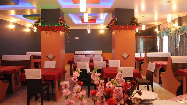 Um restaurante com várias mesas e cadeiras, algumas com toalhas brancas. Há flores decorativas e luzes fixadas na parede.