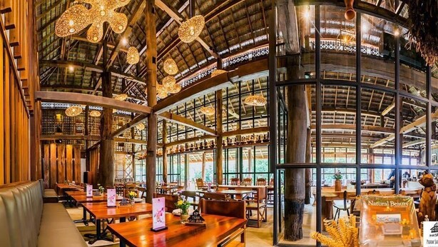 Um restaurante espaçoso com pé direito alto, vigas de madeira e amplas janelas. As mesas estão organizadas com cardápios e centros de mesa.