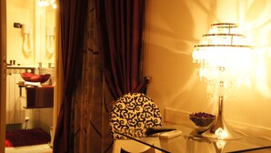 Quarto de hotel com mesa de vidro, cadeira estampada, lustre e banheiro visível ao fundo.