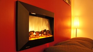 Lareira embutida na parede com fogo visível, uma luminária e uma cama com almofadas.
