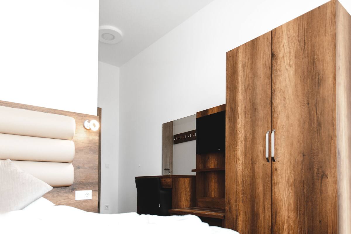 Quarto com guarda-roupa de madeira, cama com roupa de cama branca e mesa de cabeceira com abajur.