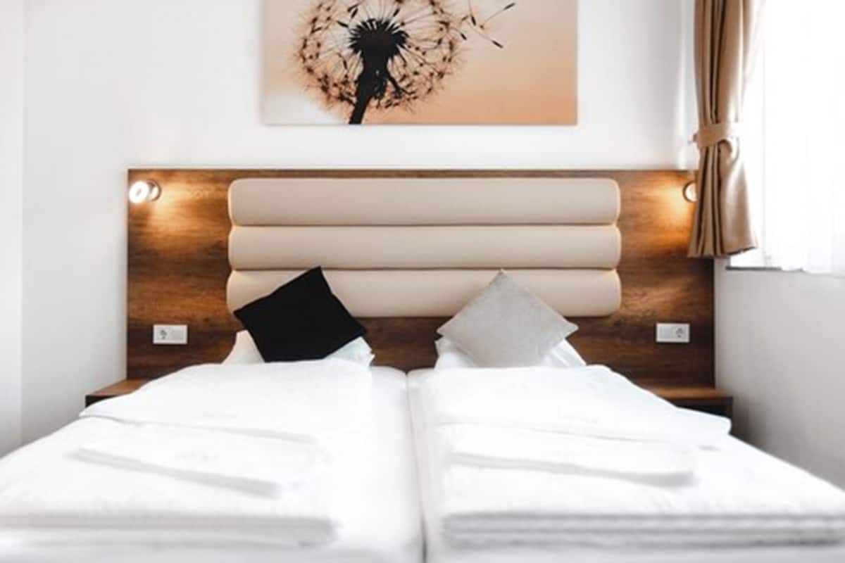 Uma cama de casal com lençóis brancos, cabeceira de madeira e uma obra de arte na parede com um dente-de-leão.