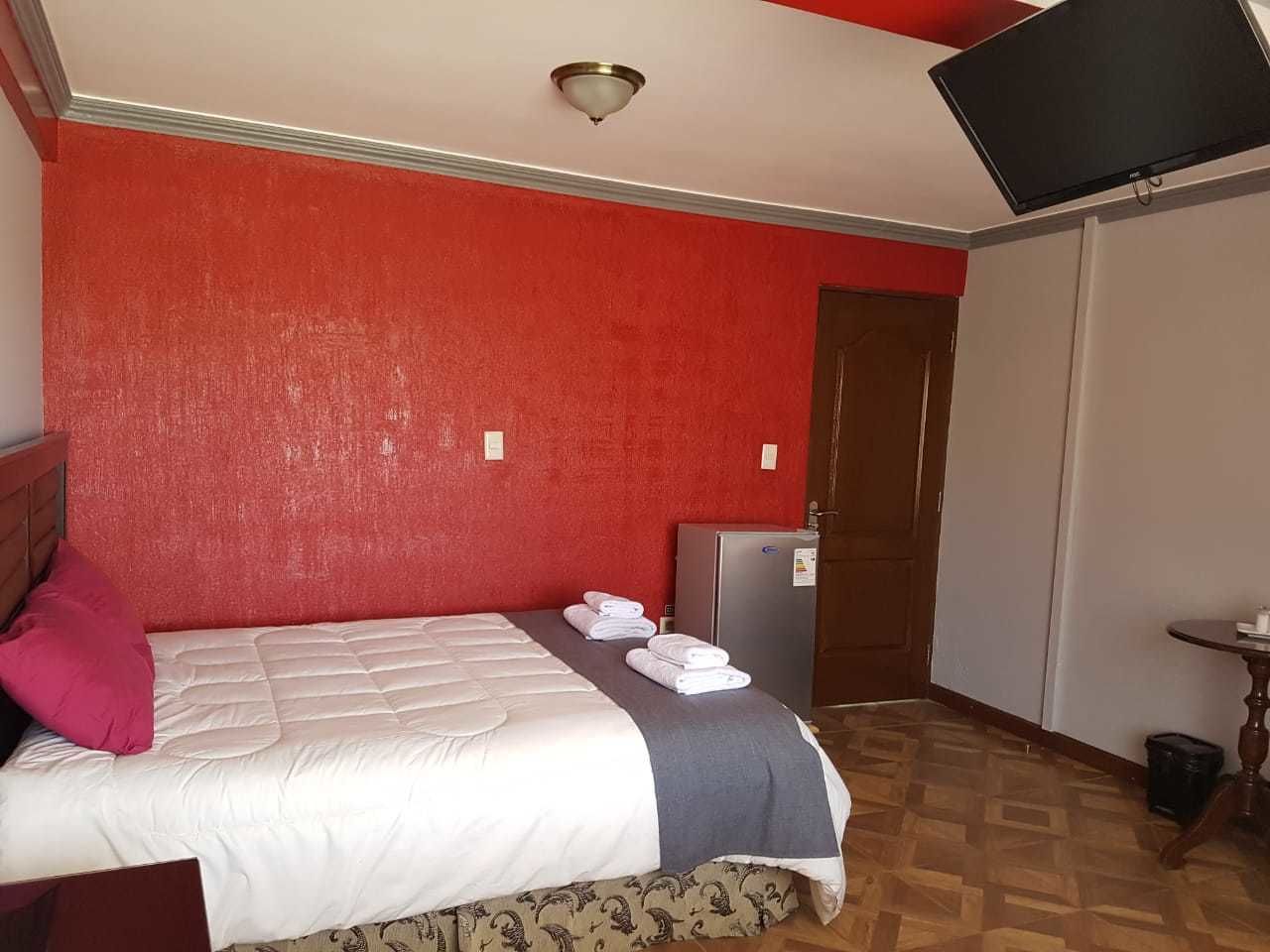 Quarto de hotel com uma cama, televisão presa na parede, um frigorífico e uma mesa pequena com uma lâmpada.