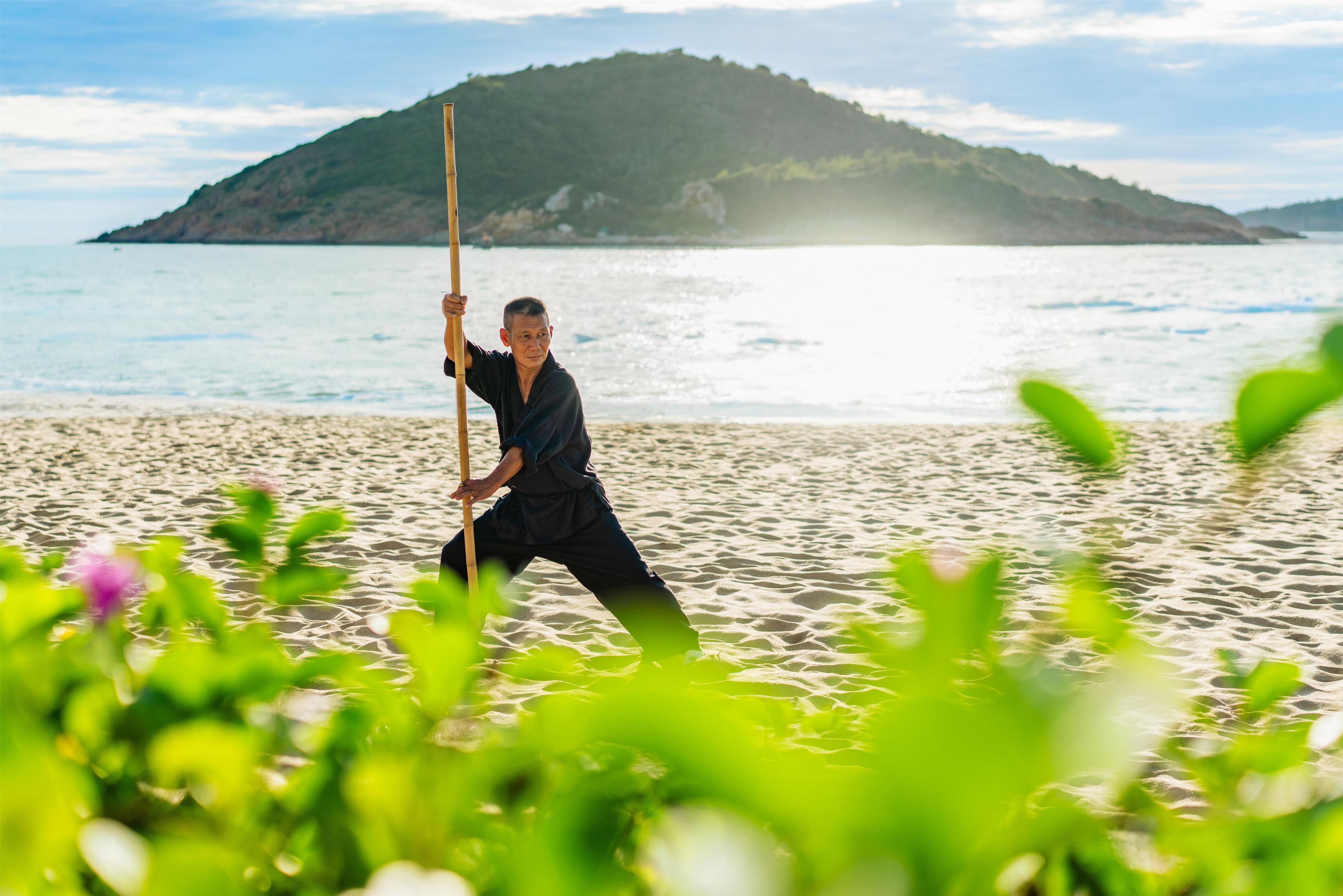 Uma pessoa praticando artes marciais numa praia arenosa, com montanhas ao fundo.