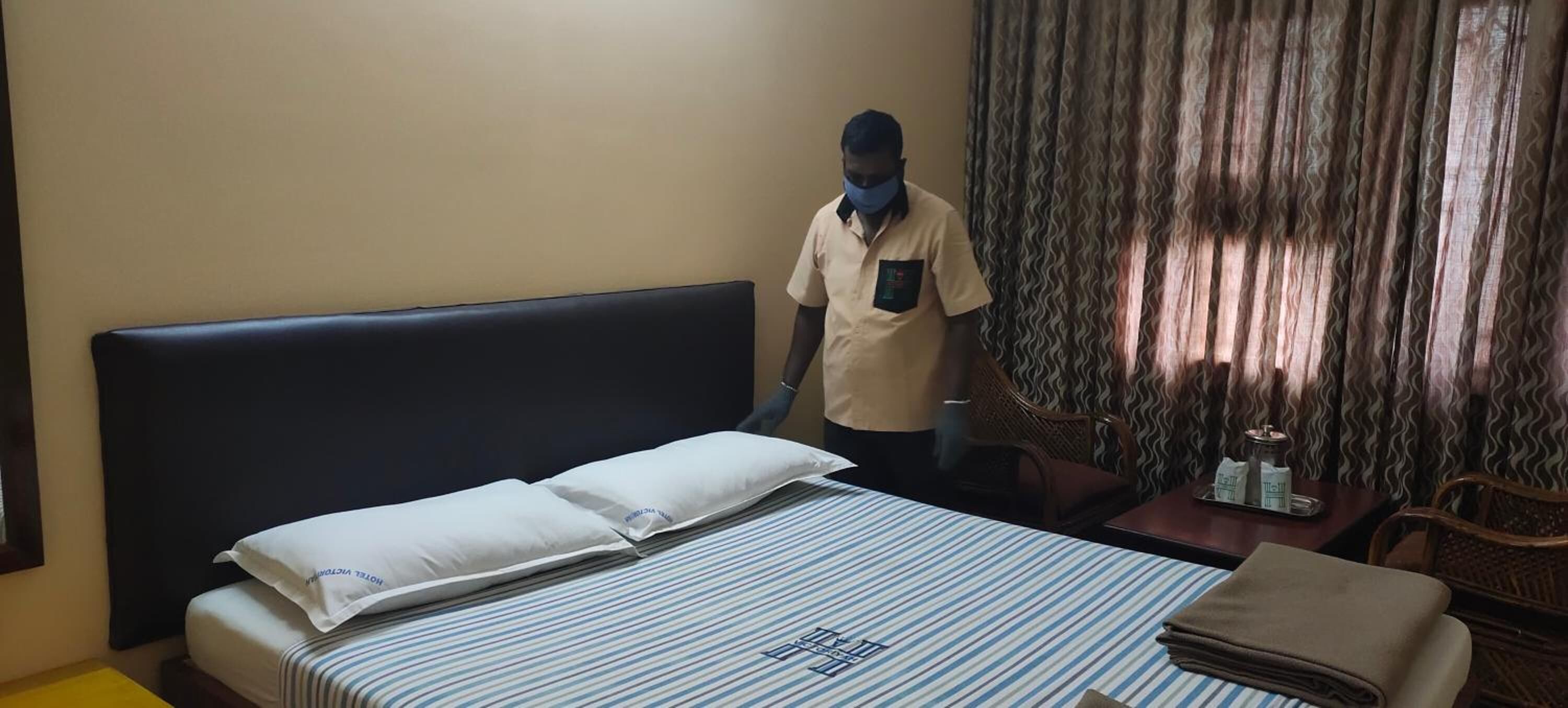 Quarto de hotel com cama, almofadas, mesa de cabeceira, cadeira e cortina.