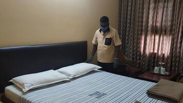 Quarto de hotel com cama, almofadas, mesa de cabeceira, cadeira e cortina.