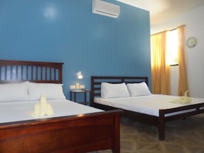 Quarto de hotel com duas camas de madeira, ar condicionado fixado na parede, pequena mesa de cabeceira com abajur e janela com cortinas.