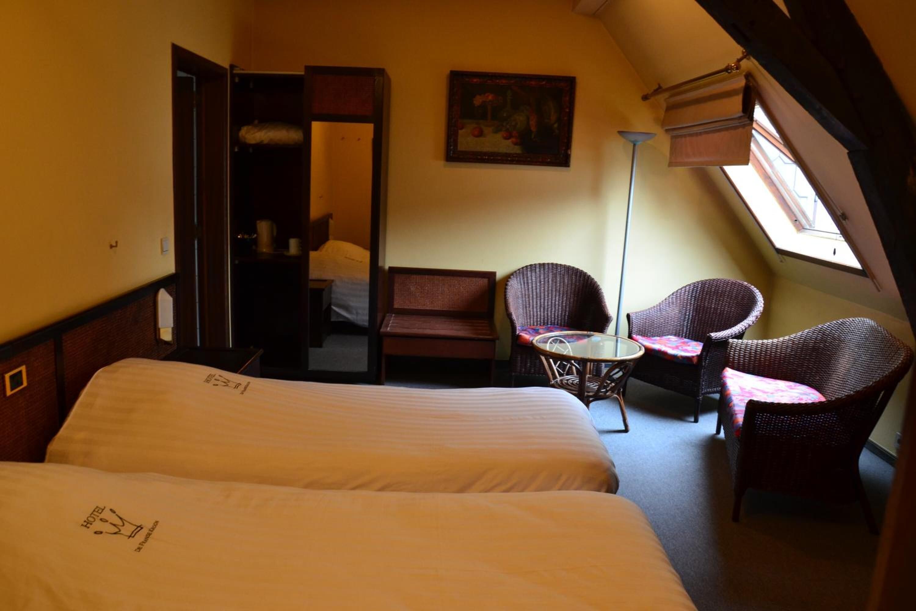 Quarto de hotel com duas camas, uma mesa pequena, cadeiras de vime, um quadro na parede e um claraboia.