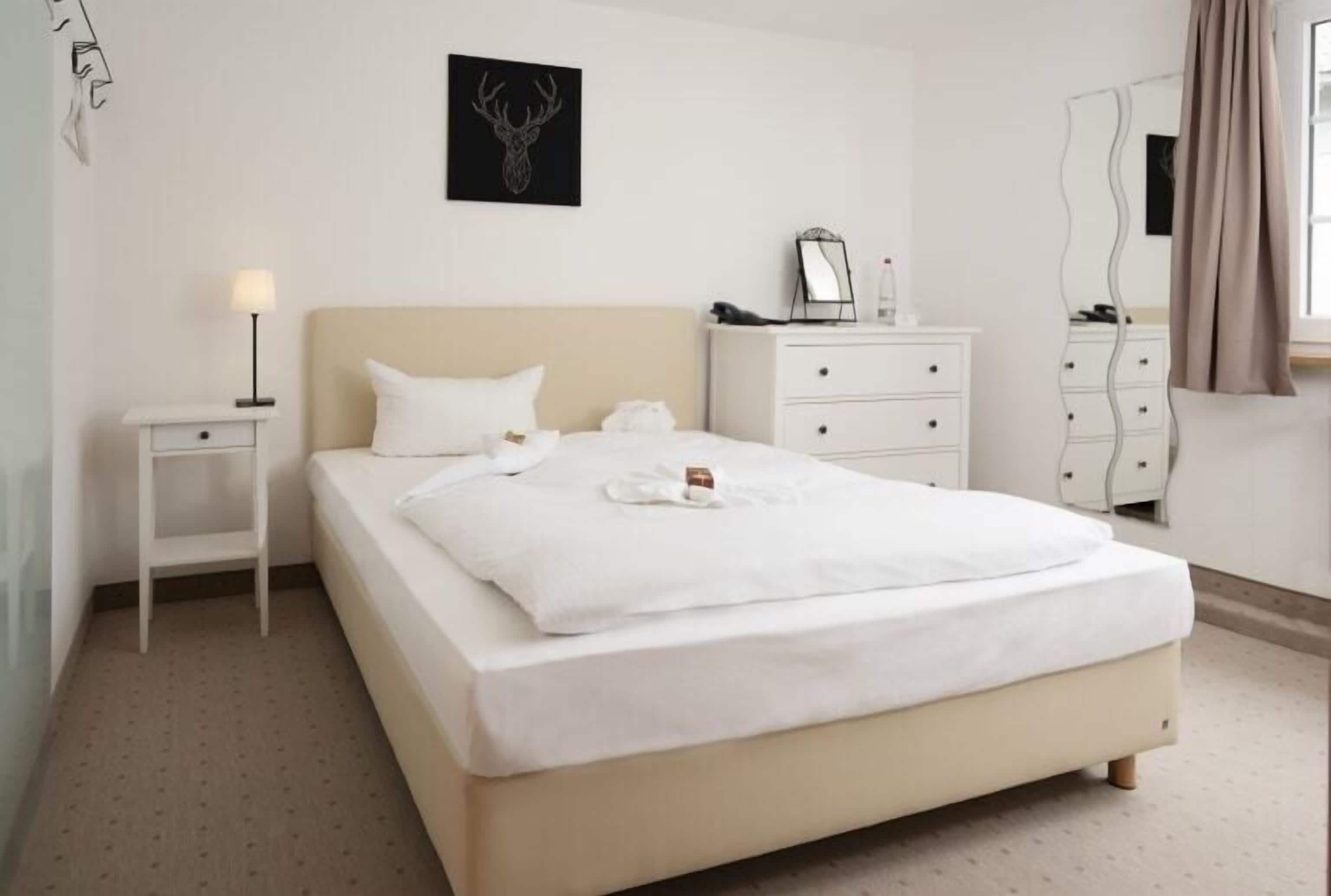 Cama bem arrumada com roupa de cama branca e um pequeno objeto decorativo. Tem uma mesa de cabeceira com luminária e espelho. O quarto tem um design simples e limpo, com esquema de cores em branco.