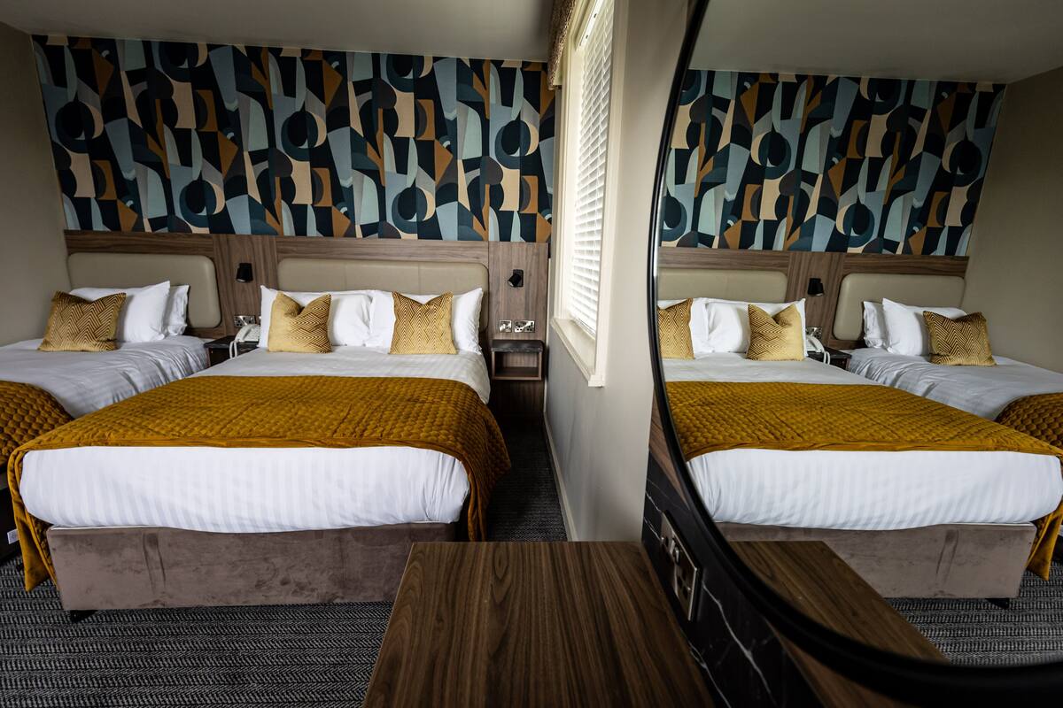 Quarto de hotel com duas camas, cada uma com roupa de cama branca e almofadas com padrão dourado, cabeceira de madeira e parede com padrão geométrico.