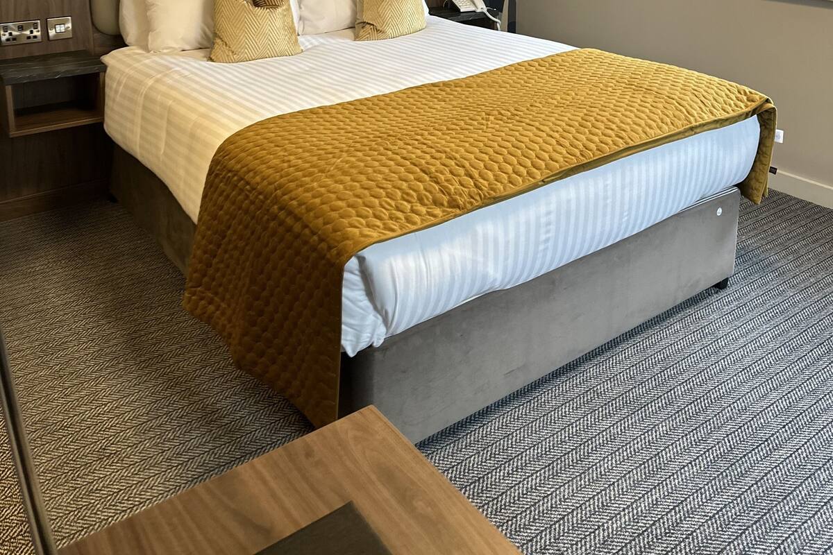 Quarto de hotel com uma cama grande, cabeceira, mesa de cabeceira, telefone fixado na parede e uma janela com persianas.