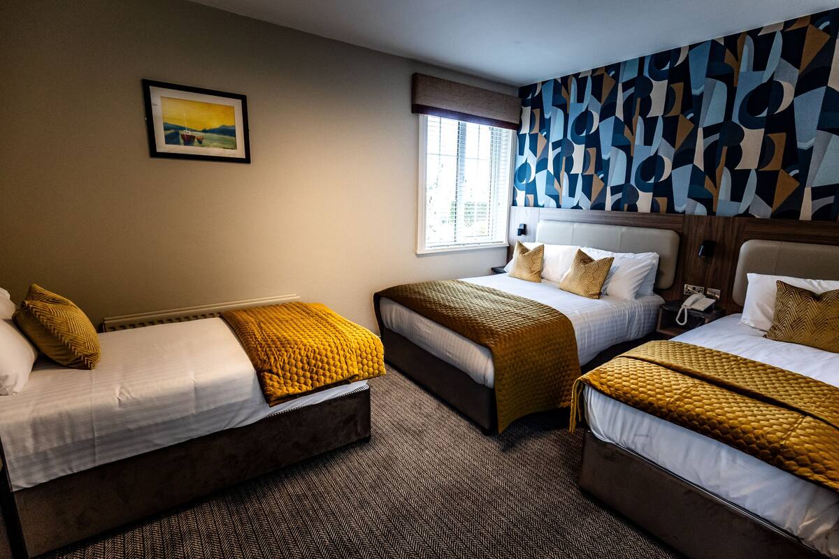 Quarto de hotel com três camas, janela com persianas, parede estampada e quadro na parede.