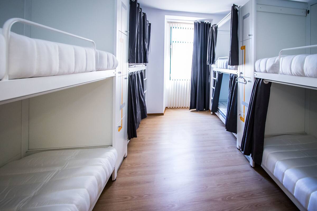 Um corredor com beliches, piso de madeira e uma janela com cortinas.
